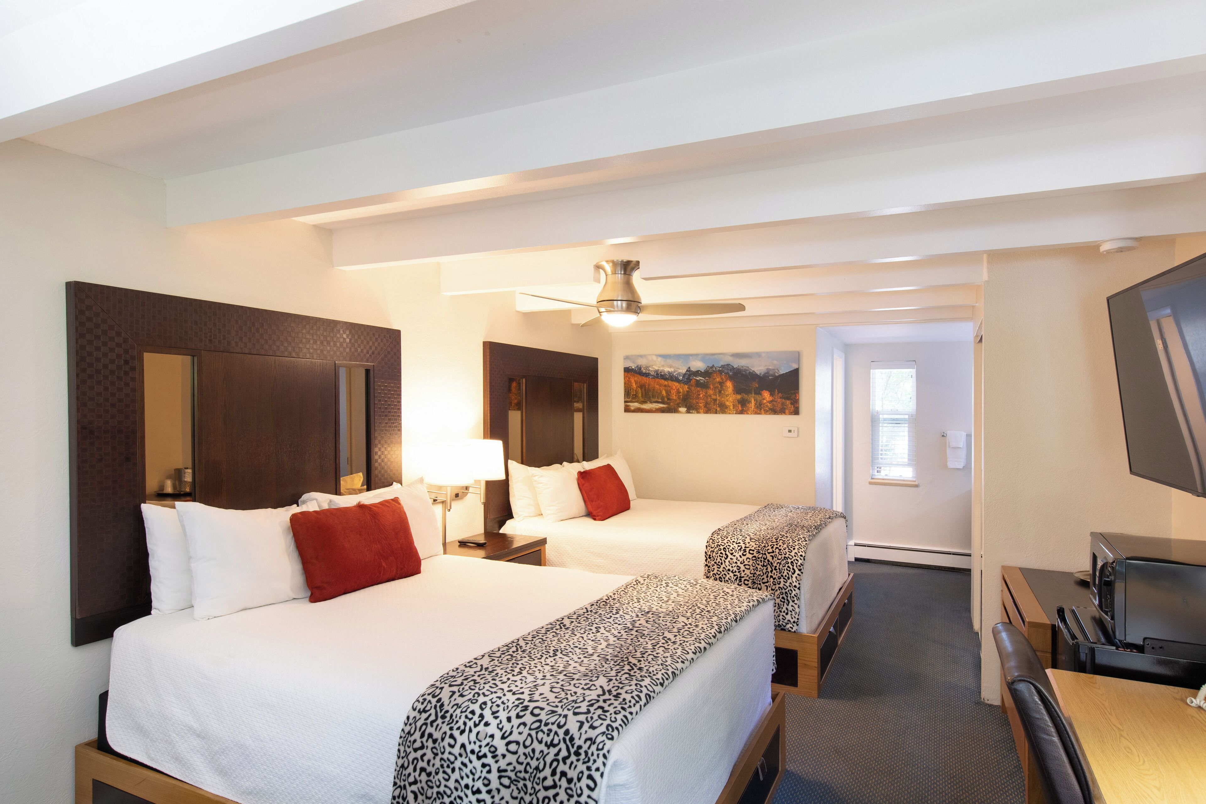 Quarto de hotel com duas camas, uma escrivaninha, televisão e ventoinha no teto.