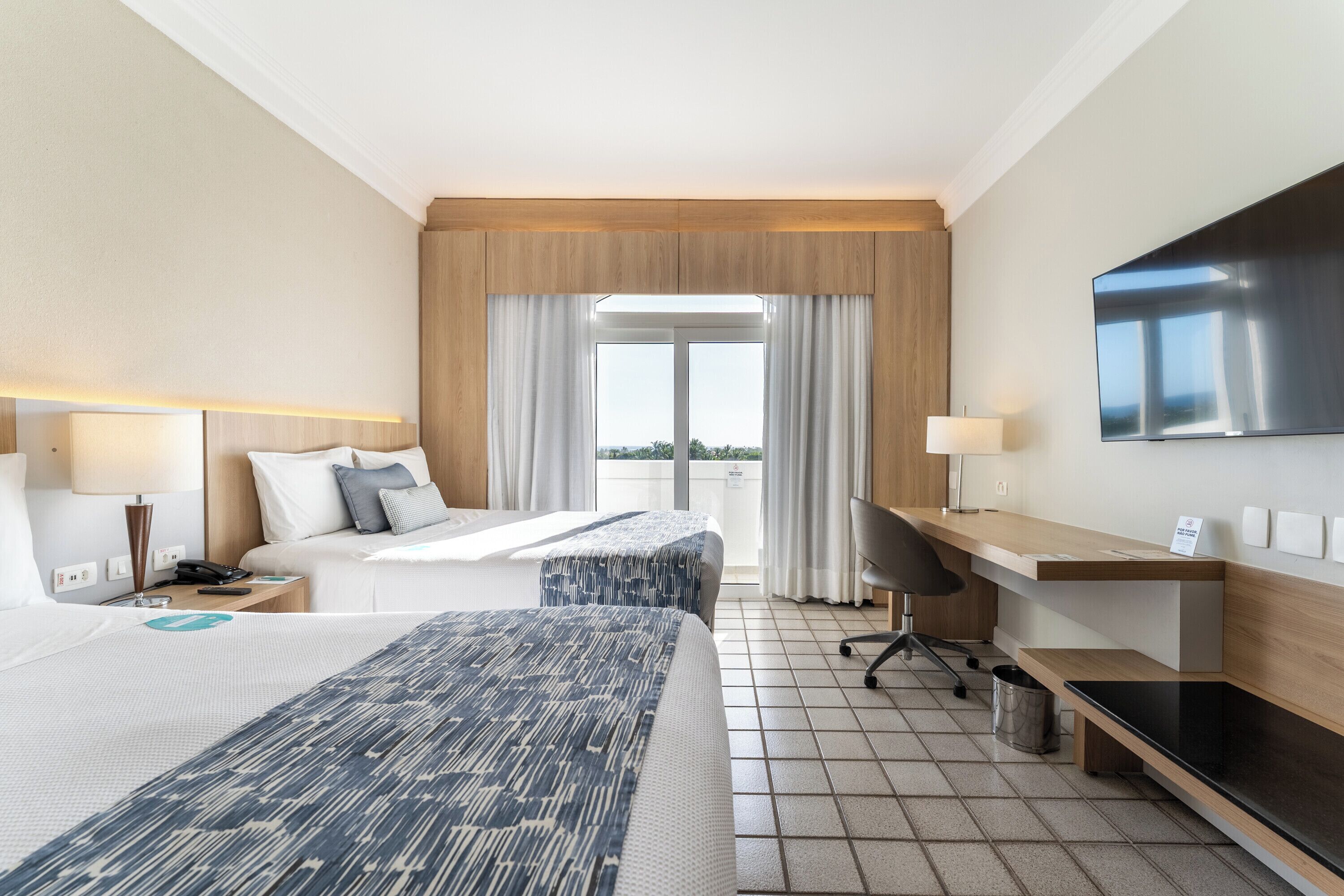 Quarto de hotel com duas camas, uma escrivaninha, uma cadeira, uma televisão e uma janela com cortinas.