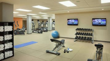 Academia com esteiras, elípticos, uma bola de exercício azul, halteres e uma garrafa de água no chão.