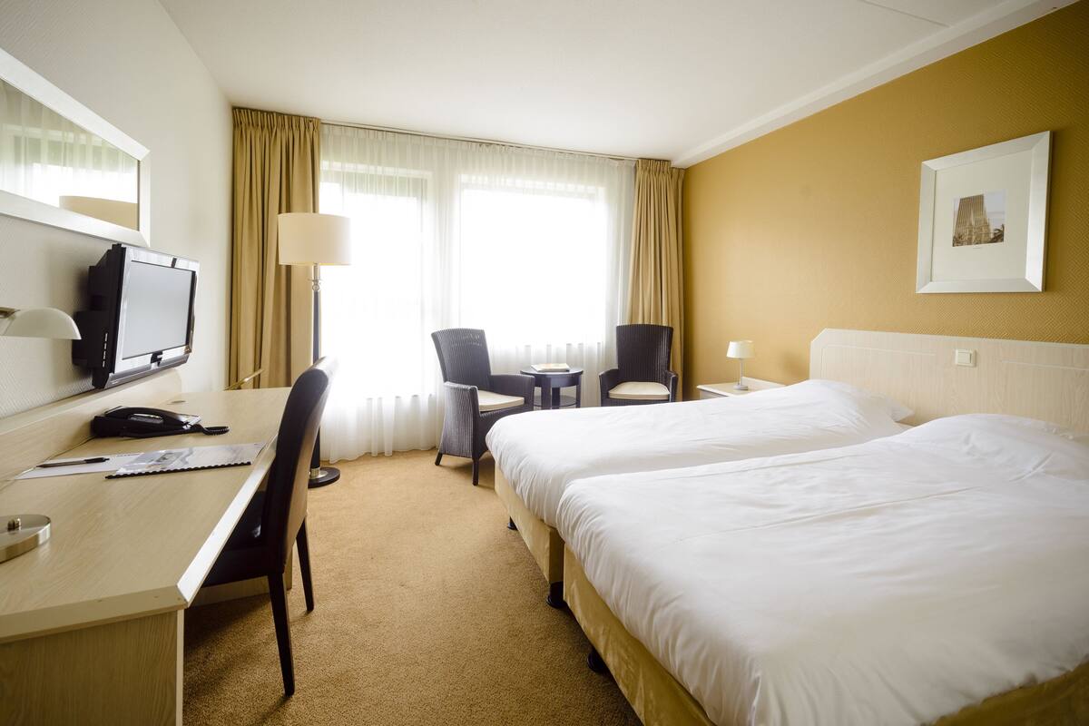 Quarto de hotel com duas camas, uma mesa de escritório com computador, uma cadeira, televisão e janela com cortinas.