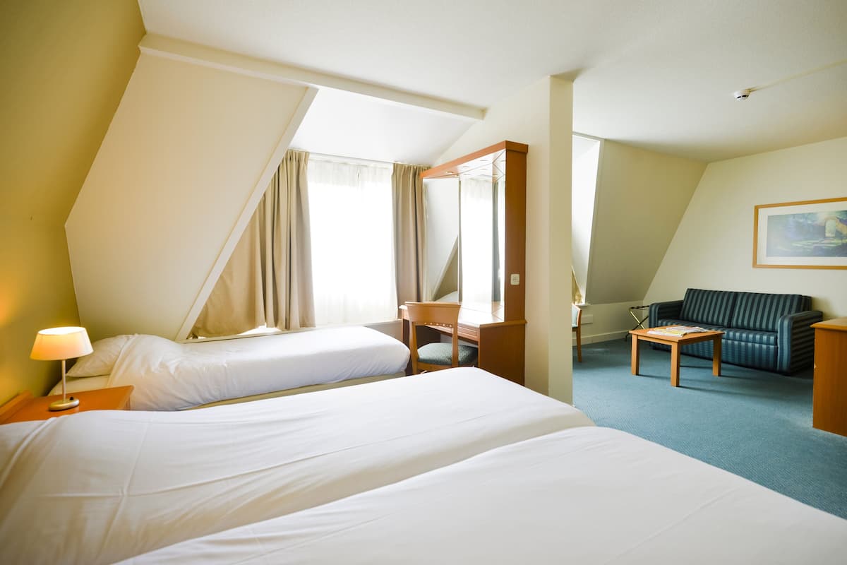 Quarto de hotel com duas camas, uma escrivaninha, uma cadeira, um sofá e uma mesa pequena.