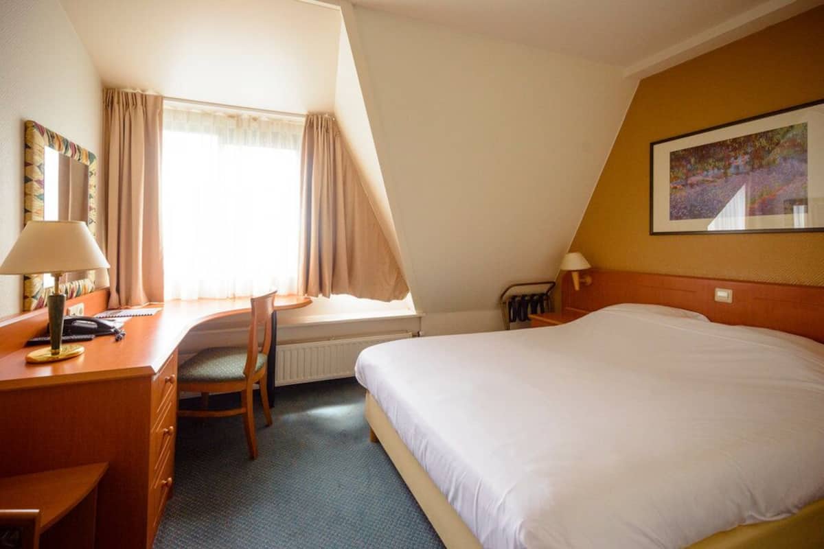 Quarto de hotel com cama, escrivaninha, cadeira e janela com cortinas.