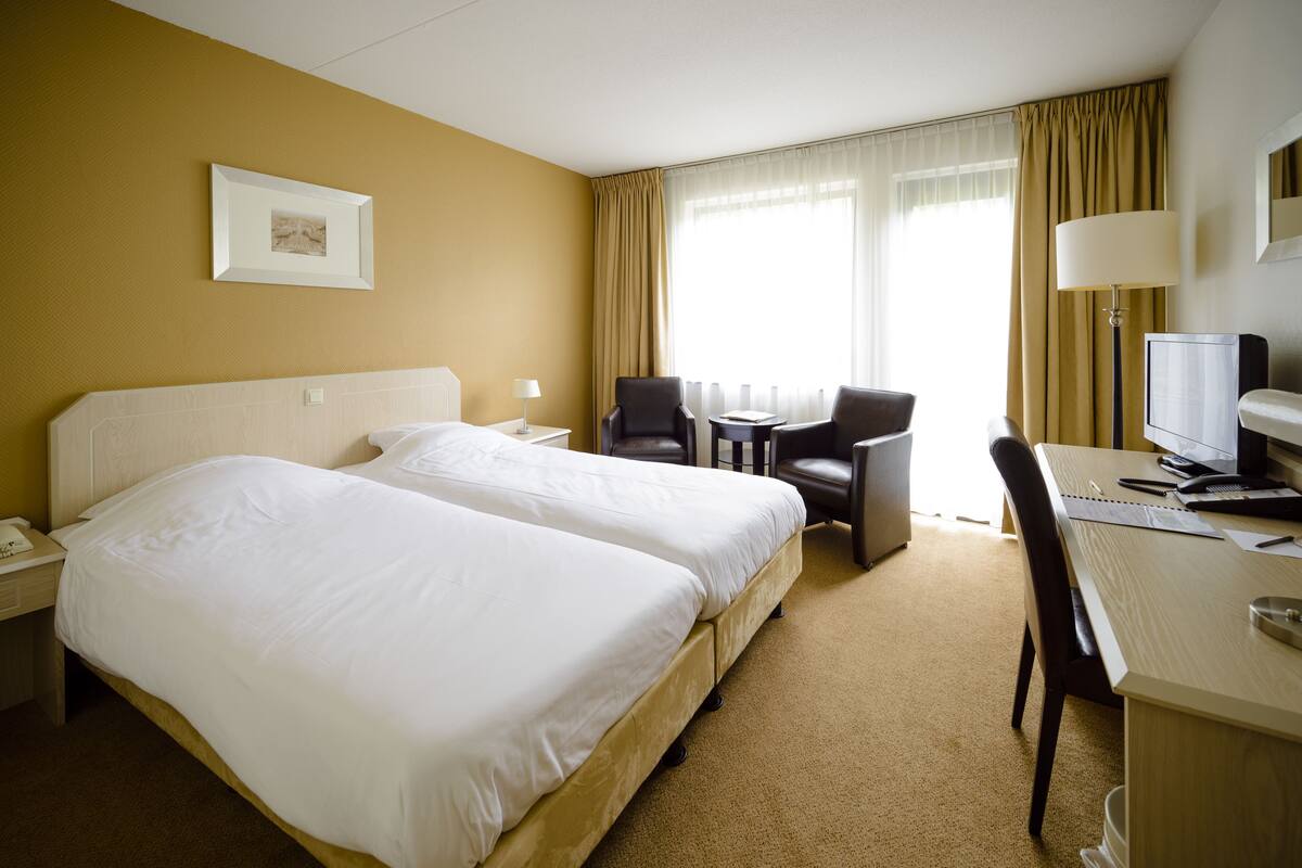 Quarto de hotel com duas camas, uma escrivaninha, uma cadeira, um abajur e uma televisão.