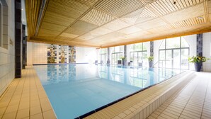 Uma grande piscina coberta com piso e teto azulejados, rodeada por janelas e plantas em vasos.