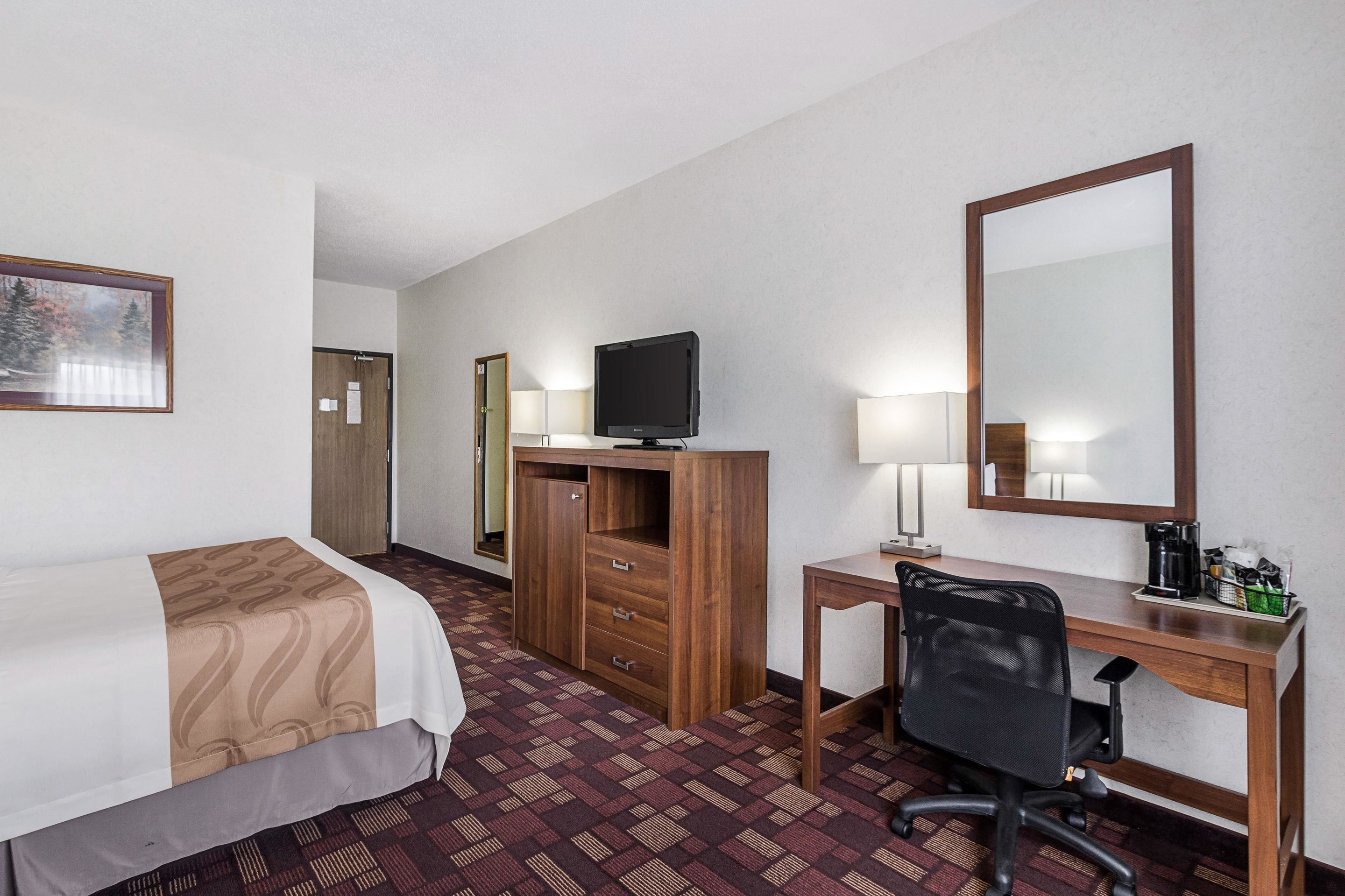 Quarto de hotel com cama, escrivaninha, cadeira, televisão, espelho e cômoda.
