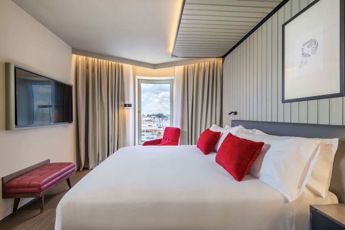 Quarto de hotel com uma cama grande, uma cadeira vermelha, um banco, uma televisão e vista para a cidade.