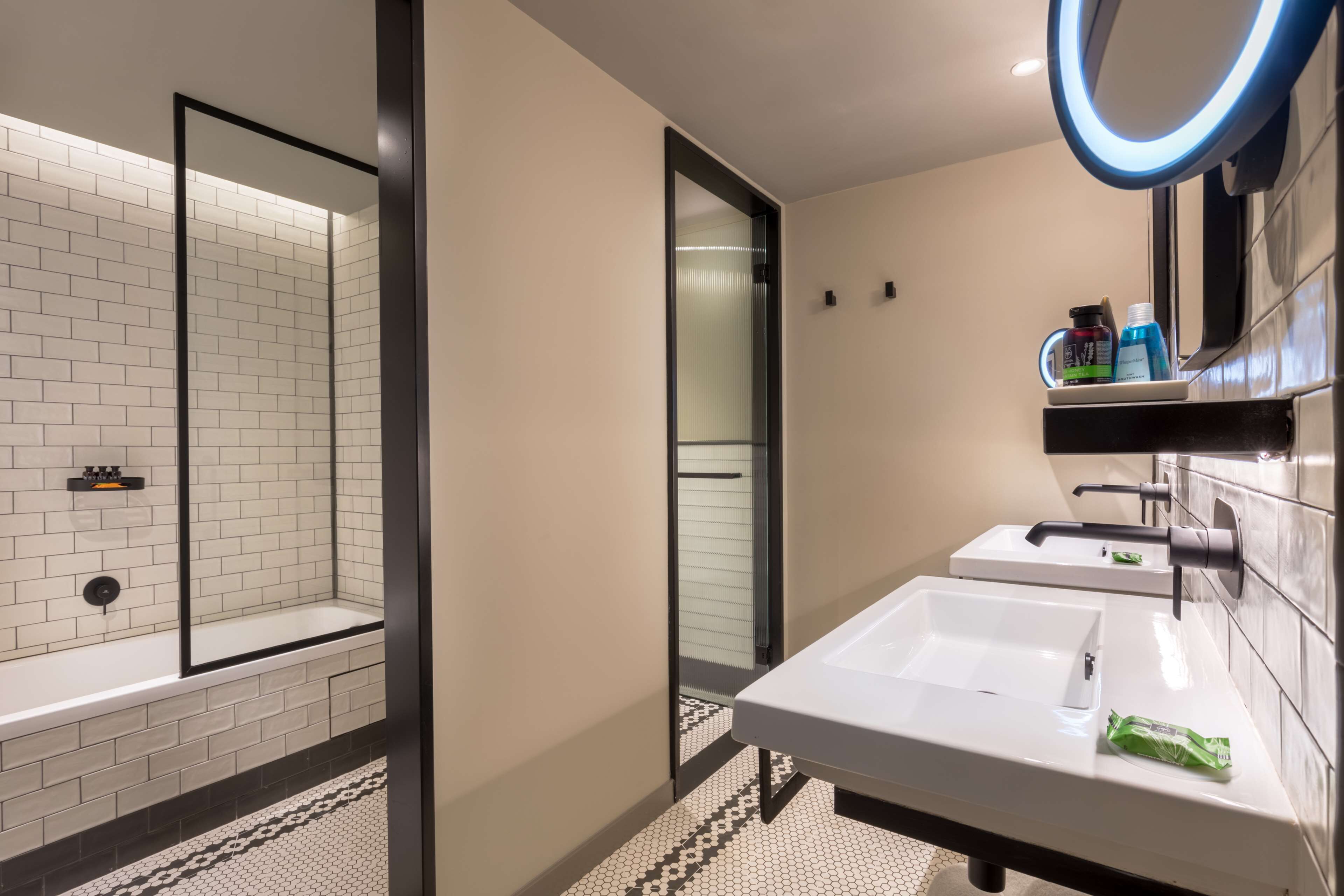 Uma casa de banho moderna com chuveiro em cabine de vidro, duas pias e um espelho com iluminação circular.