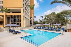 Piscina com espreguiçadeiras e guarda-sóis, cercada por palmeiras e um edifício com grandes janelas.