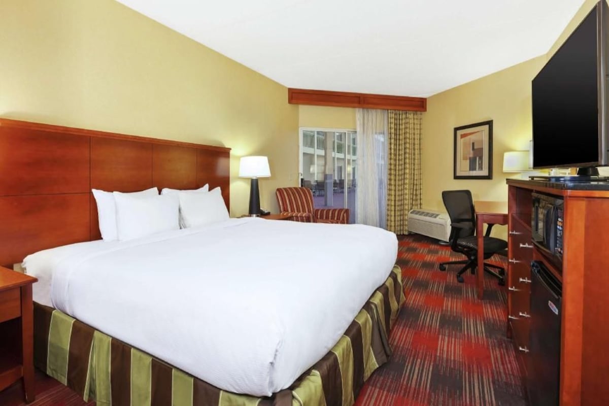 Quarto de hotel com uma cama grande, escrivaninha com computador, cadeira, televisão e janela com cortinas.