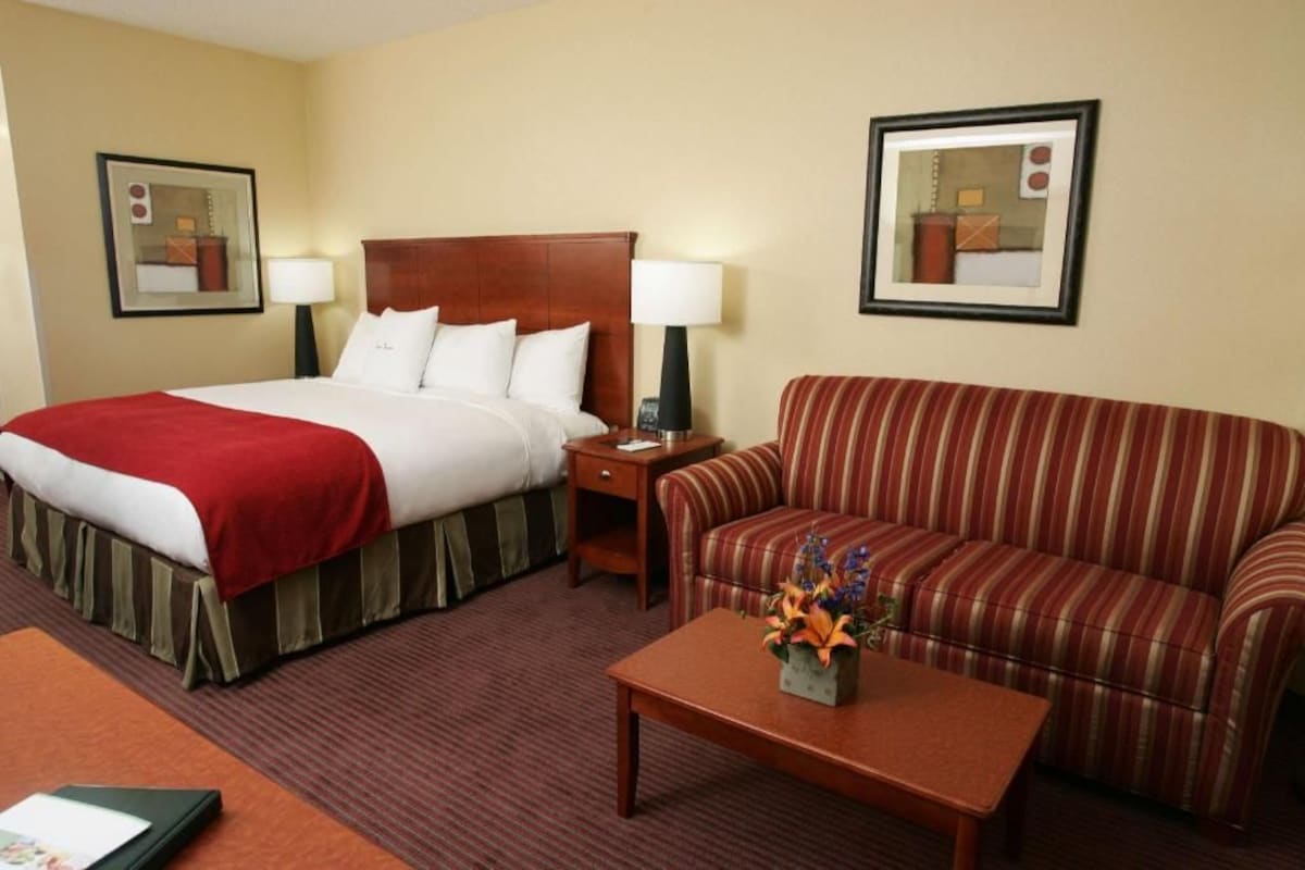Quarto de hotel com cama, sofá, mesa de cabeceira, luminária e quadro na parede.