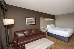 Quarto de hotel com um sofá de couro marrom, uma mesinha, uma cama com lençóis brancos, um criado-mudo, um abajur e um quadro na parede.