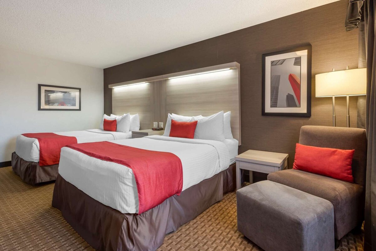 Quarto de hotel com duas camas, uma cadeira, uma mesa de cabeceira e um abajur.