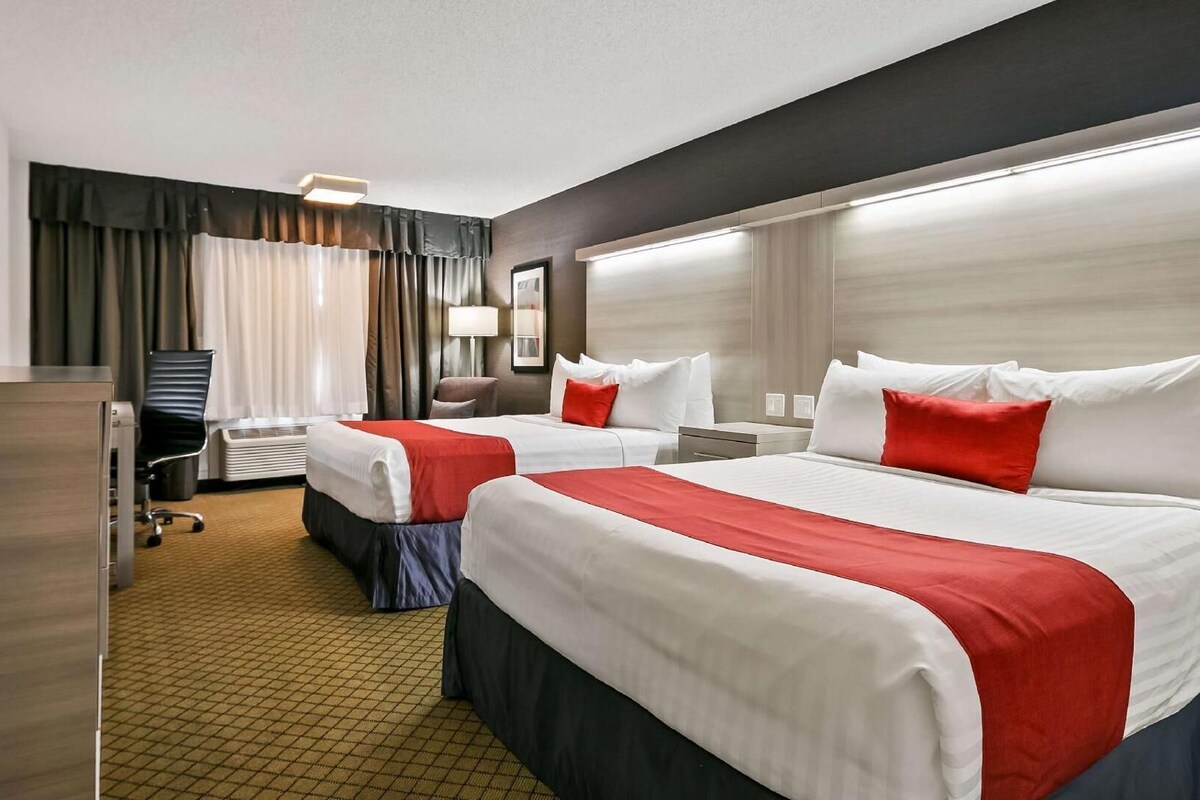 Quarto de hotel com duas camas, uma escrivaninha, uma cadeira, uma televisão e uma janela grande com cortinas.