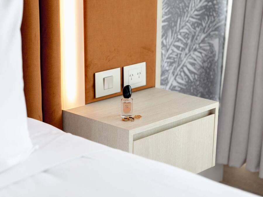 Cama bem arrumada com lençóis brancos, mesa de cabeceira de madeira com uma pequena garrafa e dois interruptores, e cortina estampada ao fundo.