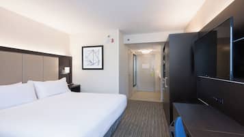 Quarto de hotel com uma cama grande, uma mesa com televisão de tela plana, um quadro na parede e uma porta que dá para um corredor.