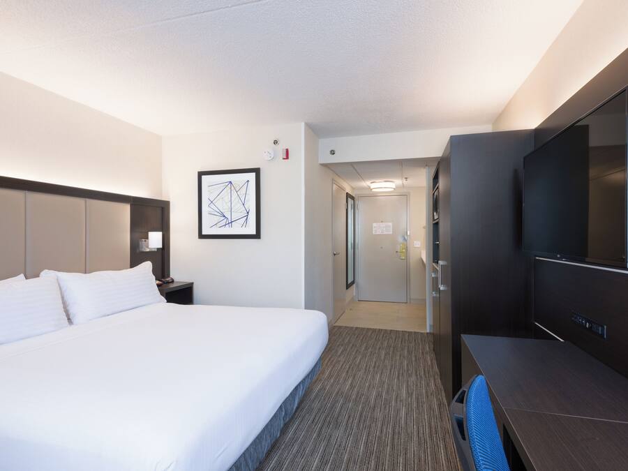 Quarto de hotel com uma cama grande, uma mesa com televisão de tela plana, um quadro na parede e uma porta que dá para um corredor.