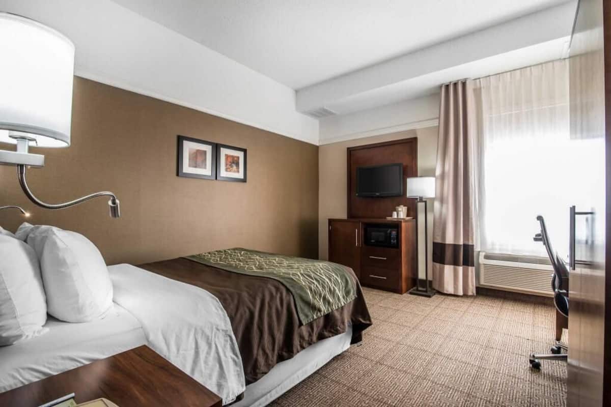 Quarto de hotel com cama, escrivaninha, televisão, cômoda e janela com cortinas.