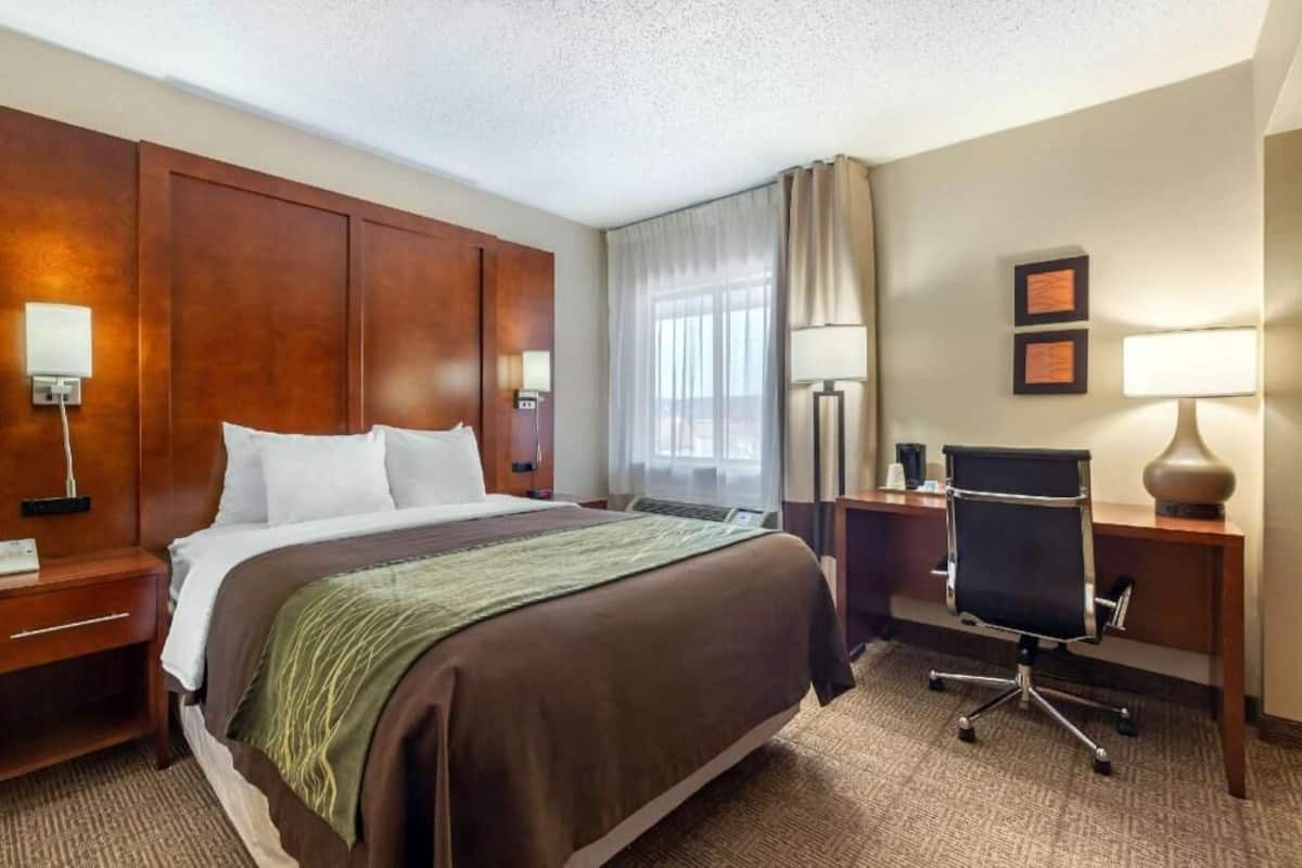 Quarto de hotel com uma cama grande, uma mesa, uma cadeira e uma janela com cortinas.