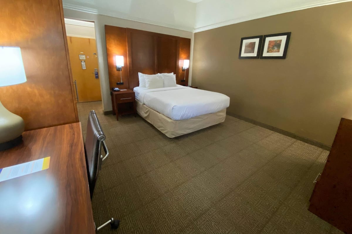 Quarto de hotel com uma cama grande, uma mesa de cabeceira de madeira, uma escrivaninha com cadeira e uma porta que dá para outro quarto.