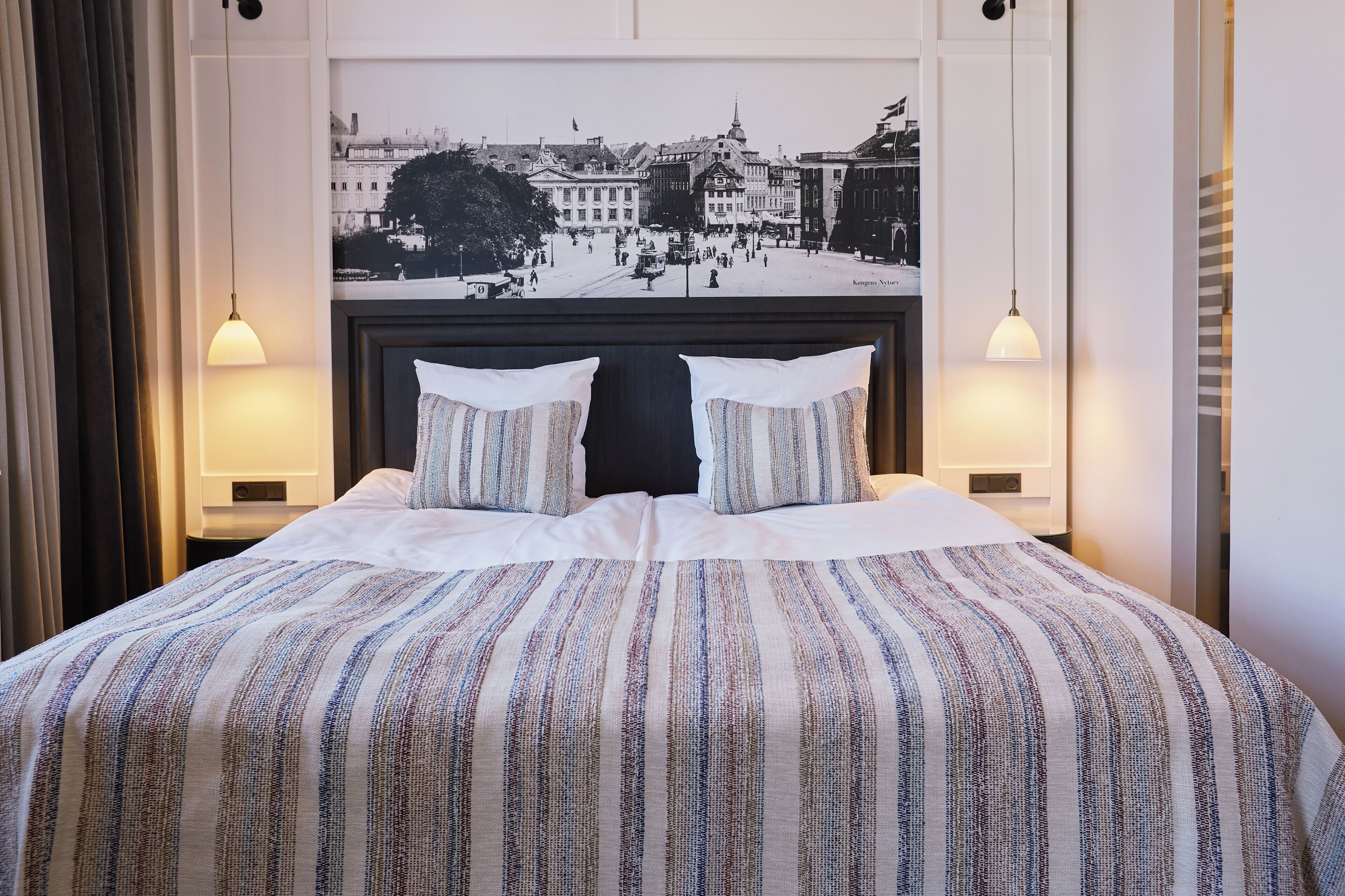 Quarto de hotel com uma cama grande, duas luminárias de cabeceira, cabeceira com estampa de paisagem urbana e vista para a cidade.