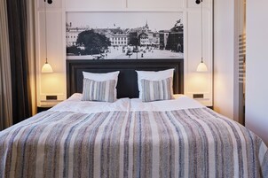 Quarto de hotel com uma cama grande, duas luminárias de cabeceira, cabeceira com estampa de paisagem urbana e vista para a cidade.