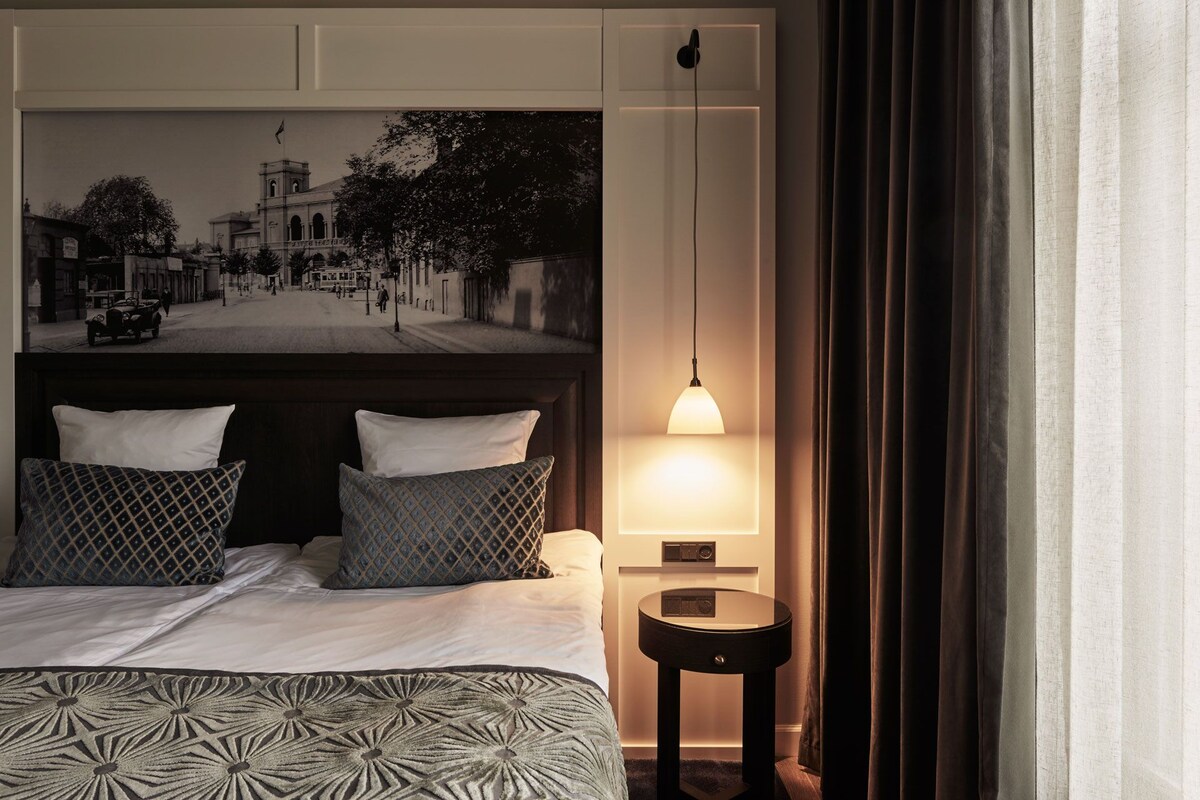 Quarto de hotel com uma cama, almofadas estampadas, uma mesinha de cabeceira, um abajur e um quadro na parede retratando uma cena de rua.
