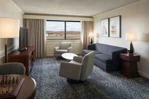 Quarto de hotel com uma janela grande, um sofá, uma cadeira, uma mesinha, uma televisão e um tabuleiro de xadrez.