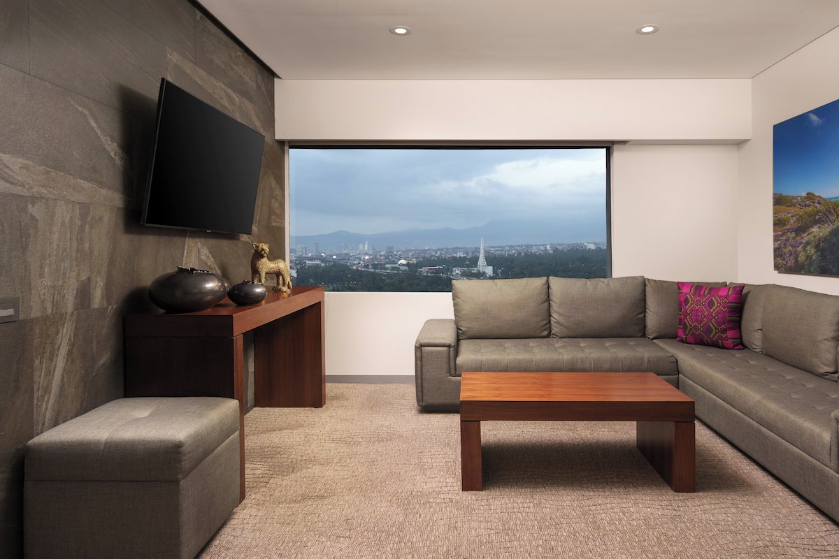 Uma sala de estar moderna com uma janela grande, uma televisão de tela plana, uma mesa de centro de madeira, um sofá cinza e um pequeno banquinho.