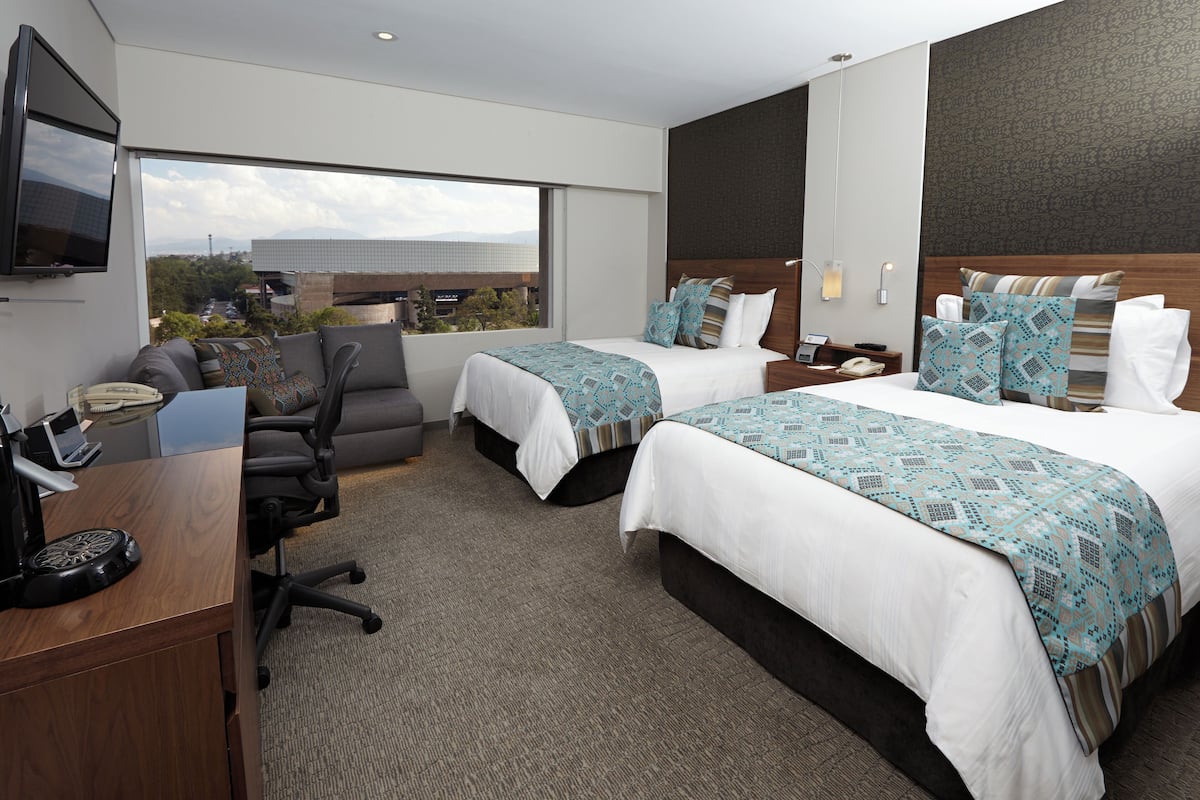 Quarto de hotel com duas camas, uma escrivaninha, uma cadeira, televisão e uma janela grande com vista para a cidade.