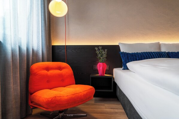 Quarto de hotel com cama, mesa de cabeceira, luminária, cadeira e janela com cortinas.