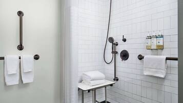 Casa de banho moderna com paredes revestidas de azulejos brancos, chão hexagonal em preto e branco, chuveiro com chuveiro manual, porta-toalhas e uma pequena mesa com toalhas dobradas.