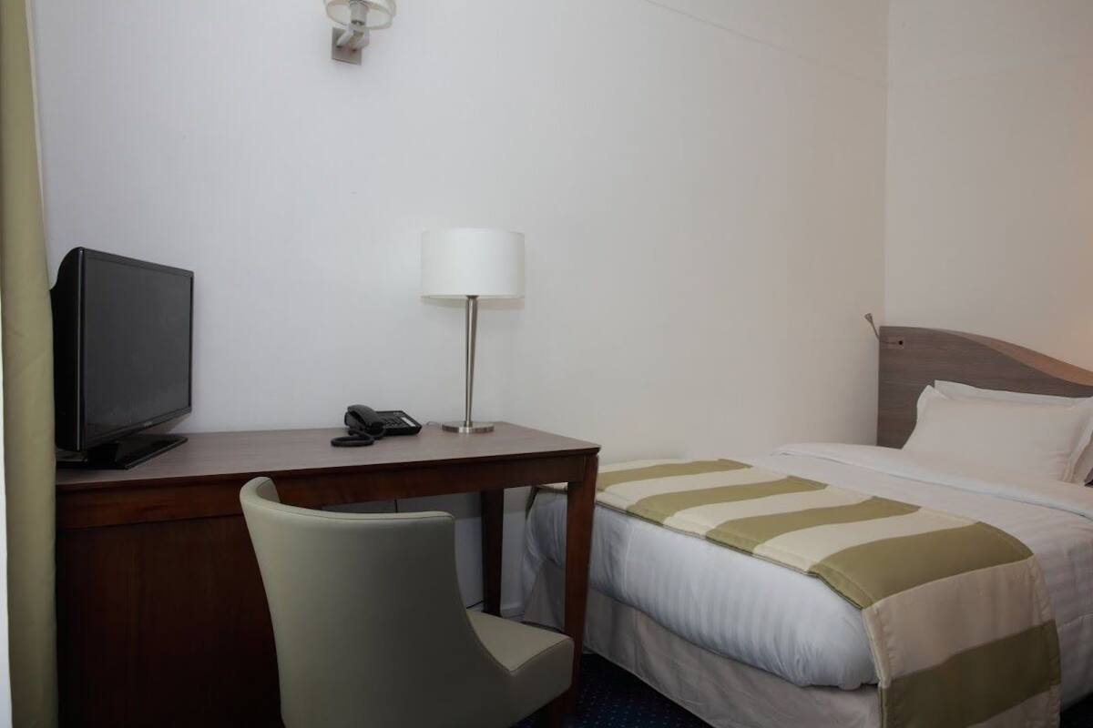Quarto de hotel com cama, escrivaninha com televisão, luminária e cadeira.