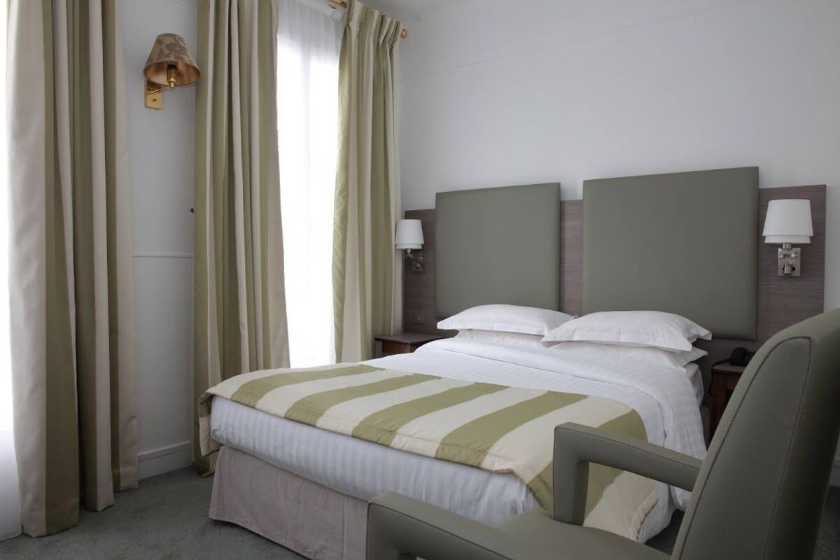 Quarto de hotel com uma cama, duas mesas de cabeceira, uma cadeira e cortinas.