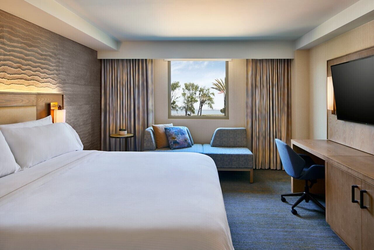 Quarto de hotel com uma cama grande, uma escrivaninha, uma cadeira, uma televisão e uma janela com vista para árvores e o céu.