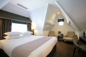 Quarto de hotel com uma cama grande, duas poltronas, uma mesa pequena e uma janela com cortinas.