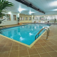 Uma piscina interior com água azul cristalina, rodeada por paredes azulejadas e um teto com grelhas de ar condicionado.