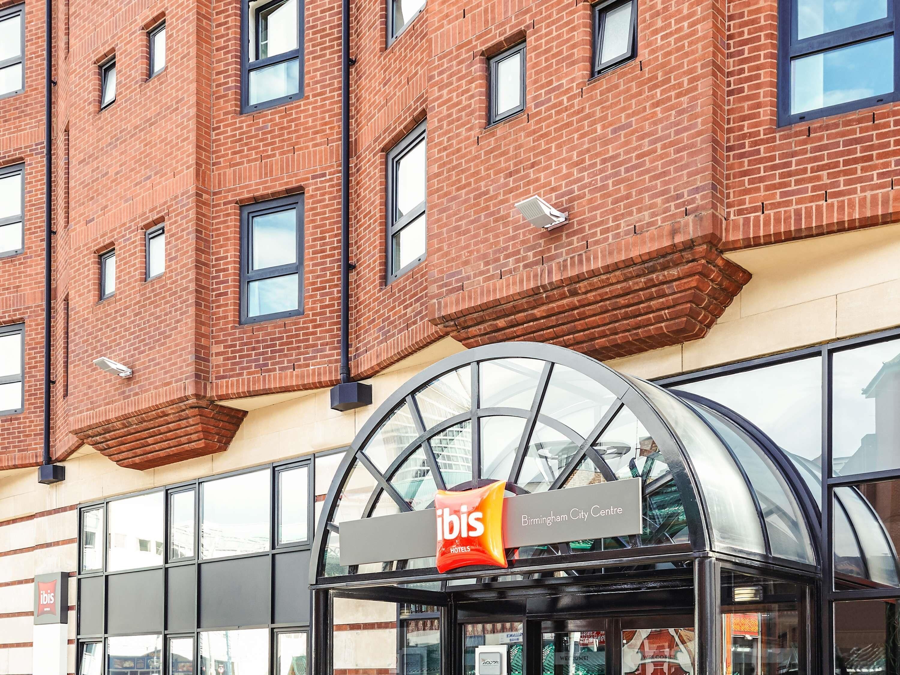 Entrada de hotel com toldo de vidro, sinalização para 'ibis Hotels' e fachada de um edifício de tijolos.