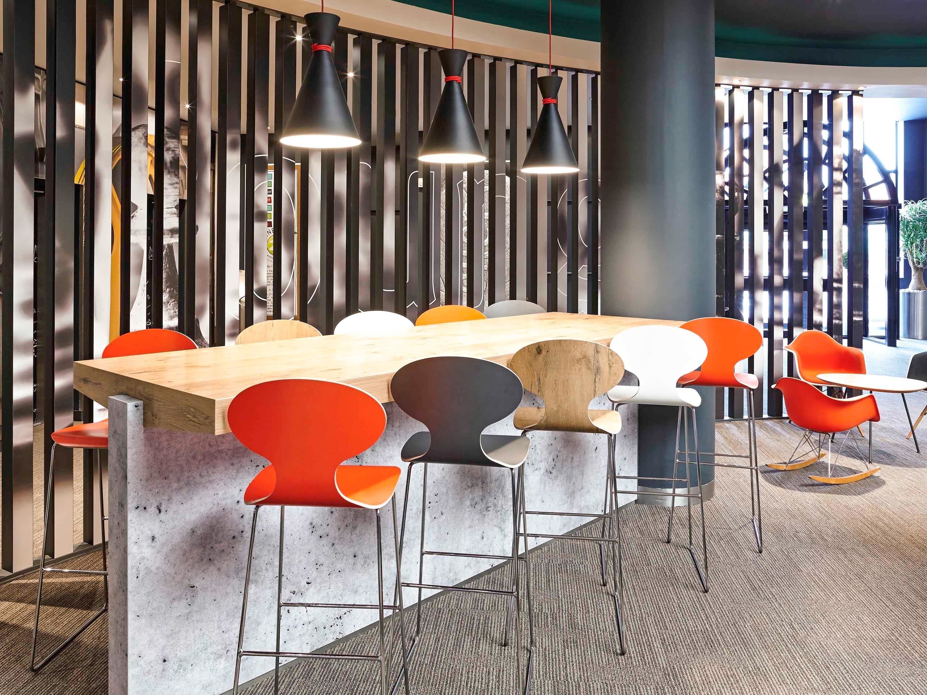 Uma sala de jantar moderna com uma mesa de madeira comprida, cadeiras coloridas e iluminação pendente.