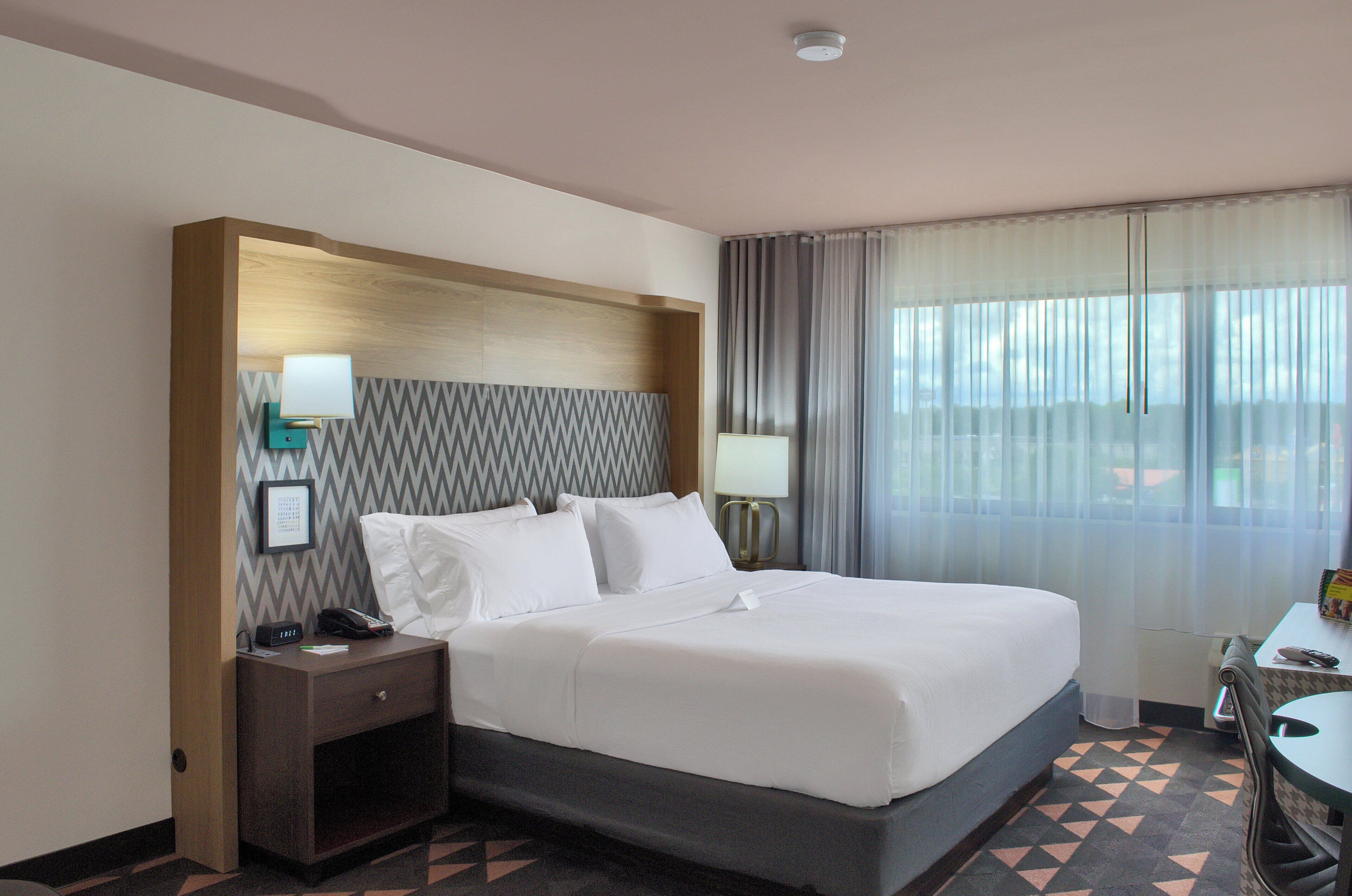 Quarto de hotel com cama, mesa de cabeceira, luminária e vista para o exterior pela janela.