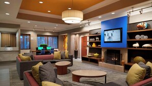 Um hall de hotel moderno com área de estar, lareira e televisão exibindo um jogo esportivo.