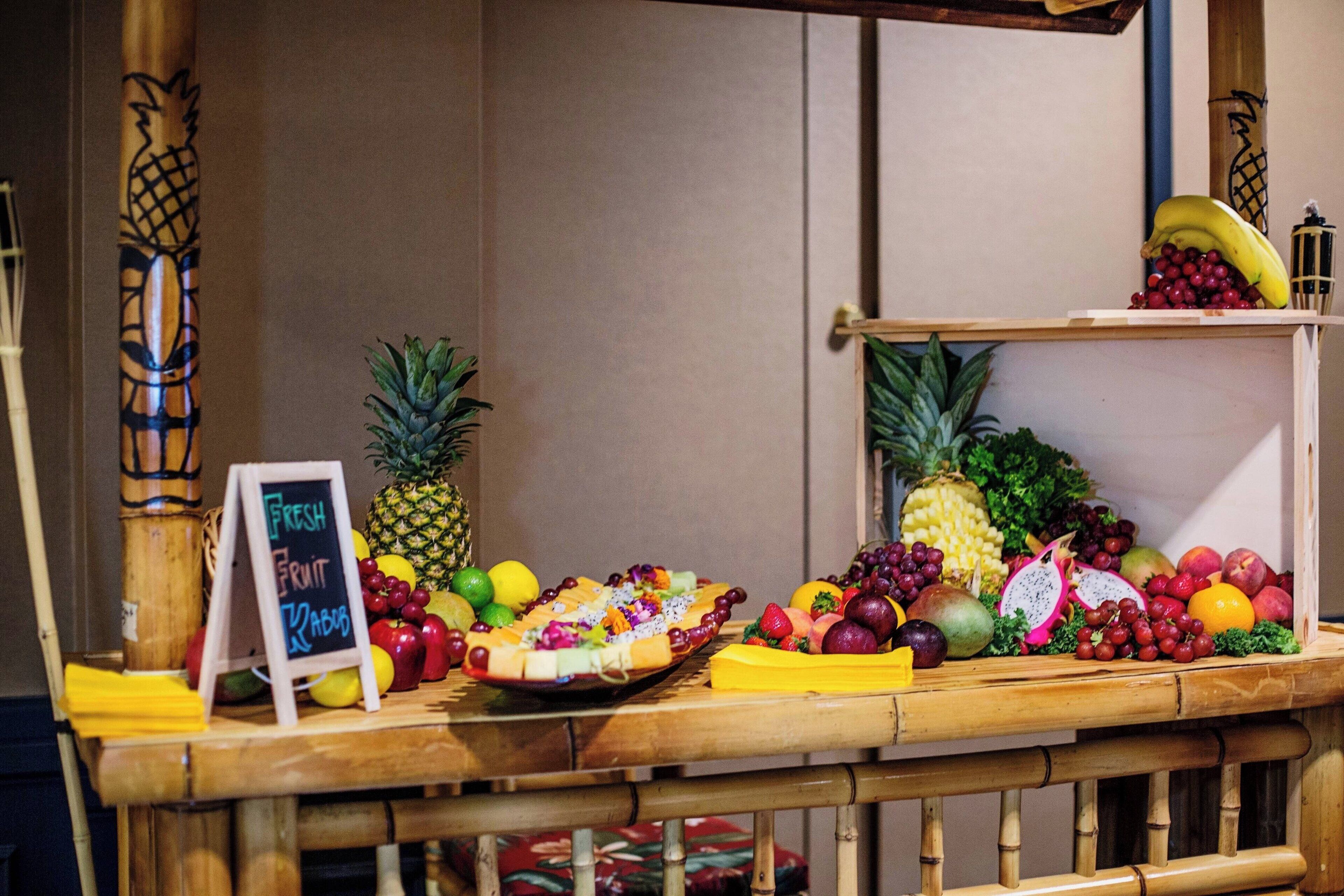 Exposição de frutas com uma placa indicando 'Frutas Frescas' e diversas frutas como abacaxi, uvas e maçãs.
