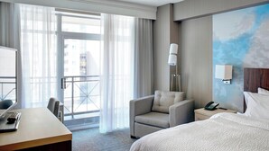 Quarto de hotel com cama, escrivaninha, cadeira, mesa de cabeceira com telefone e janela com cortinas.