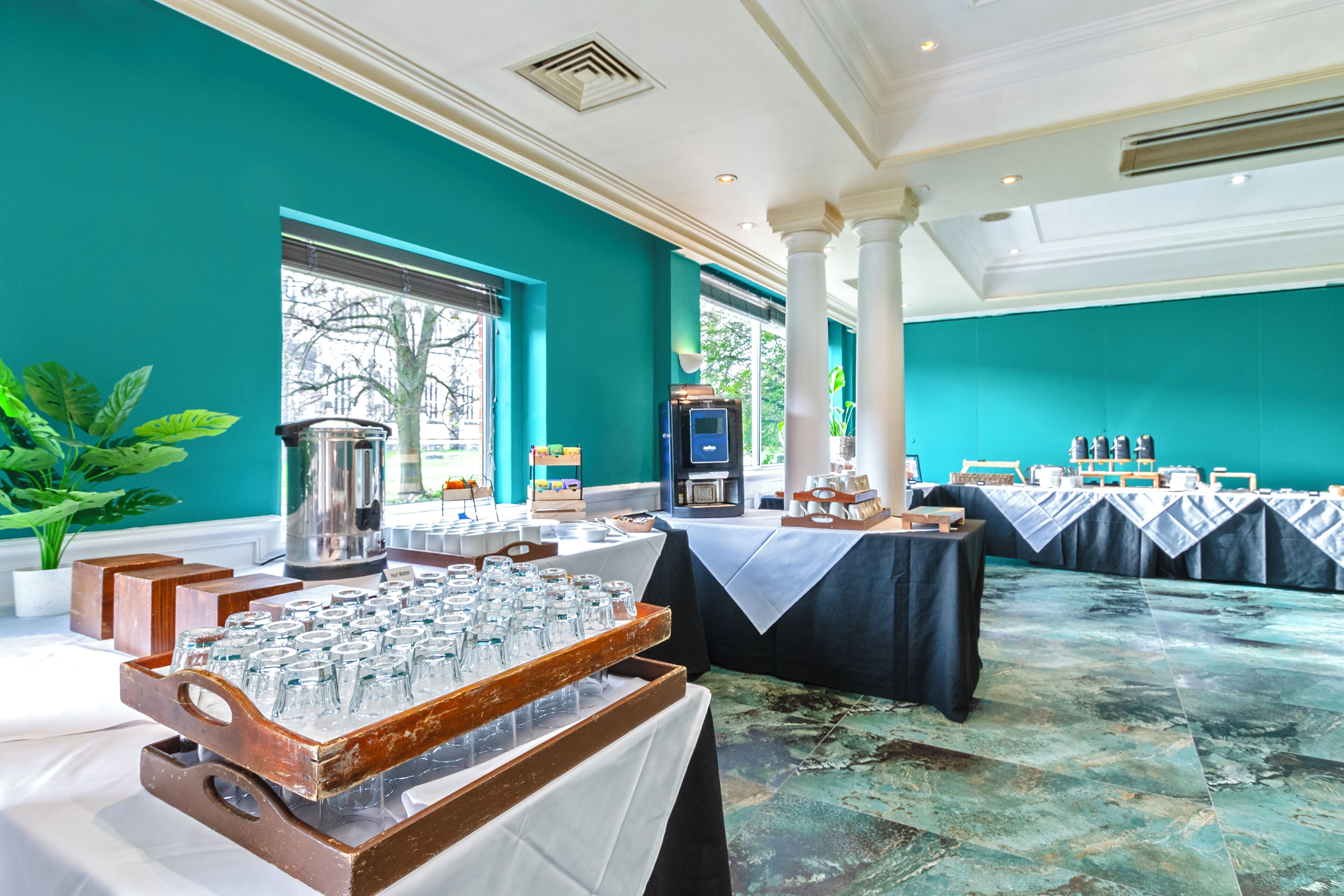 Um quarto espaçoso com paredes cor de verde-água, grandes janelas e piso de mármore. Há mesas montadas com copos e uma área de buffet.