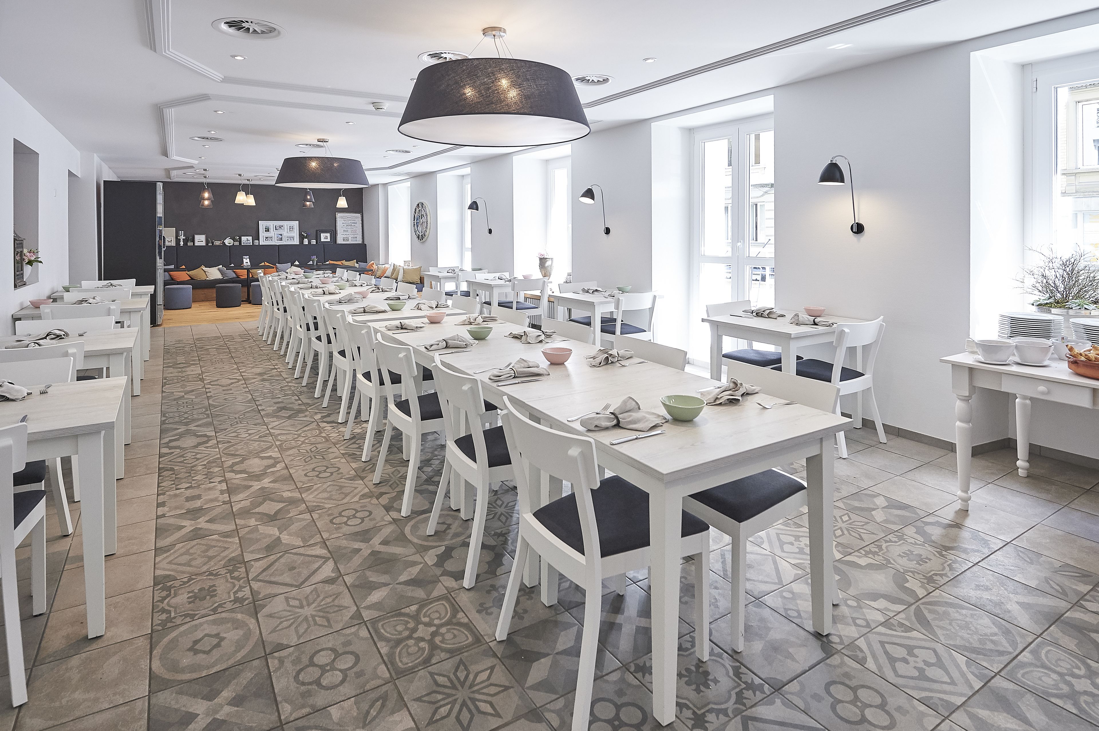 Uma sala de jantar moderna com mesas e cadeiras brancas, ladrilhos estampados no chão e iluminação pendente.