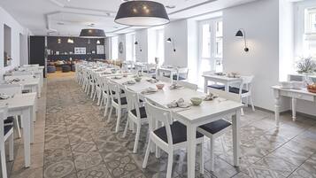 Uma sala de jantar moderna com mesas e cadeiras brancas, ladrilhos estampados no chão e iluminação pendente.