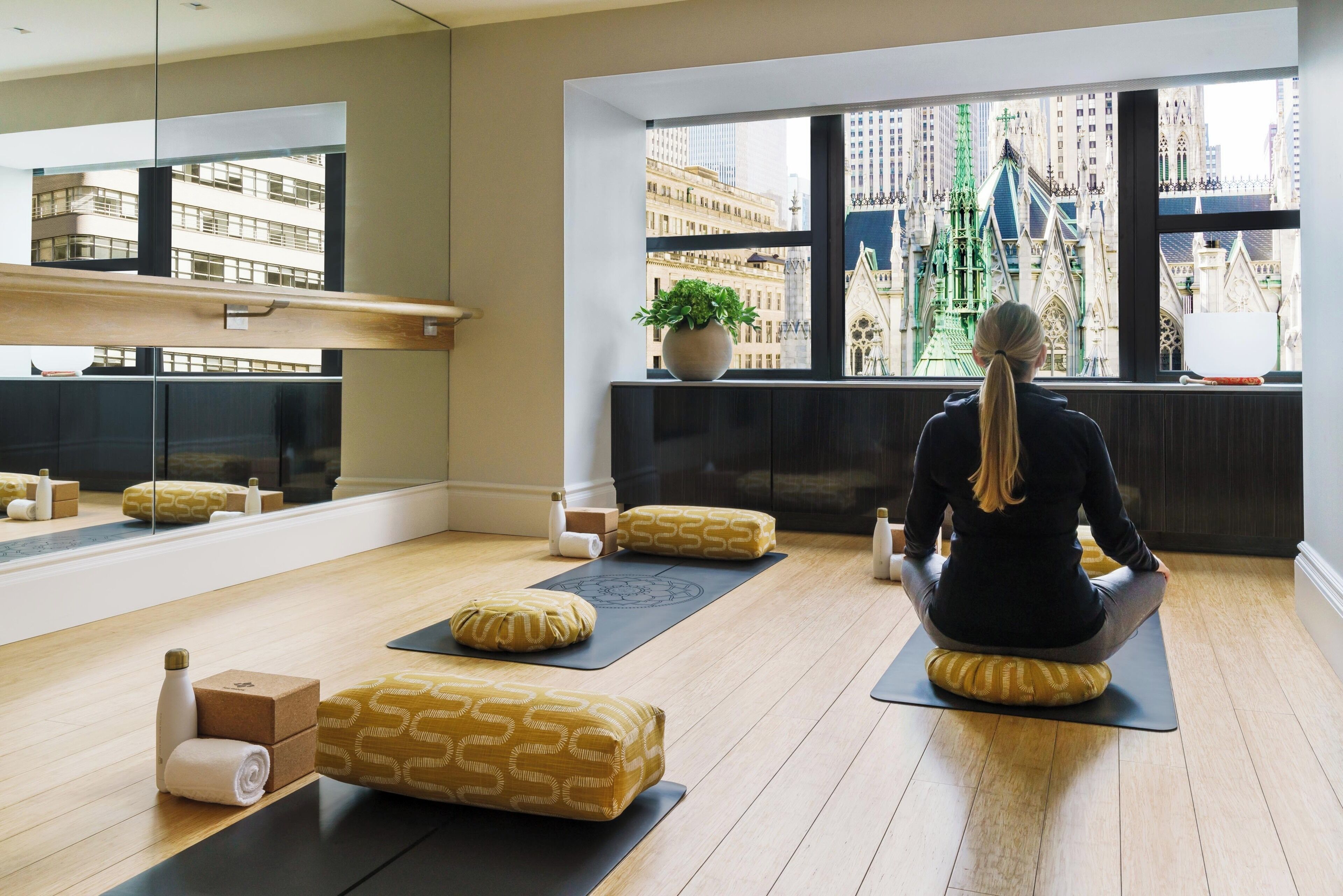 Uma sala de yoga com piso de madeira, tapetes pretos e uma grande janela com vista para um edifício histórico.