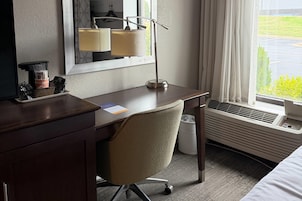 Quarto de hotel com escrivaninha, cadeira, luminária, janela com cortinas e vista para o exterior.