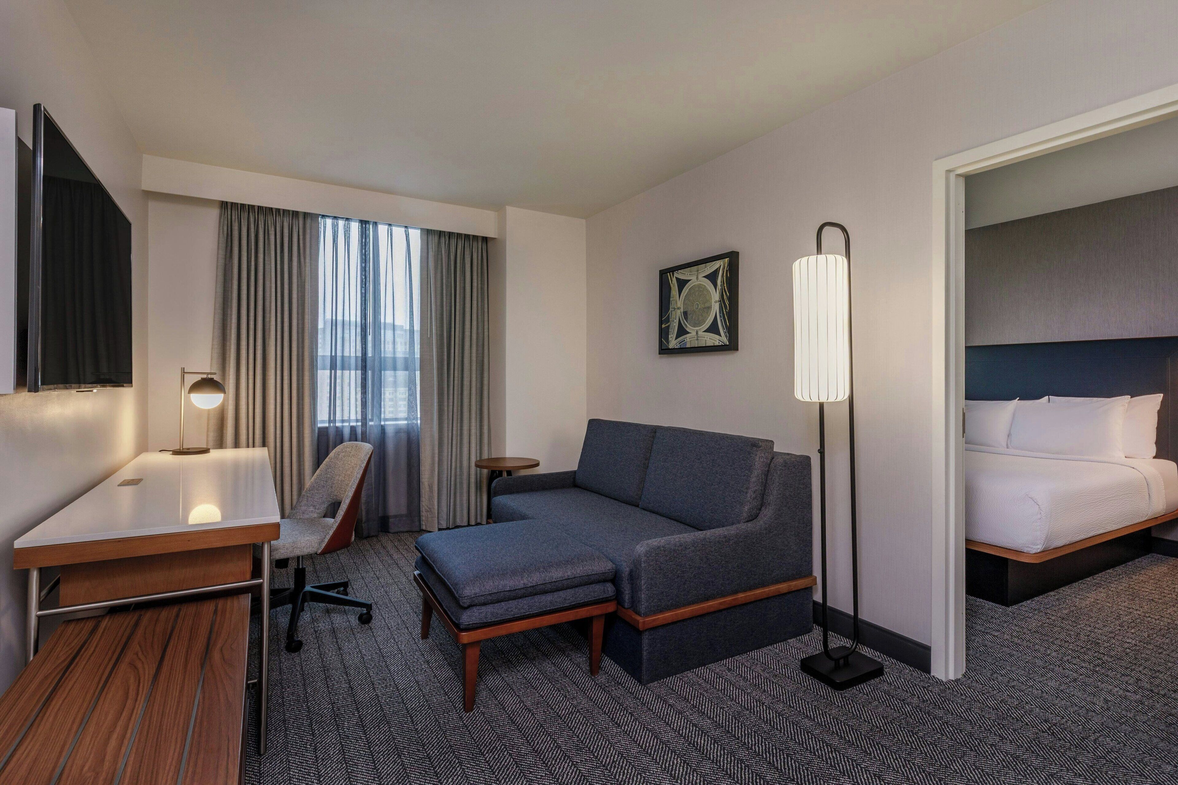 Quarto de hotel com escrivaninha, cadeira, sofá, apoio de pés, mesa de cabeceira, abajur e cama.
