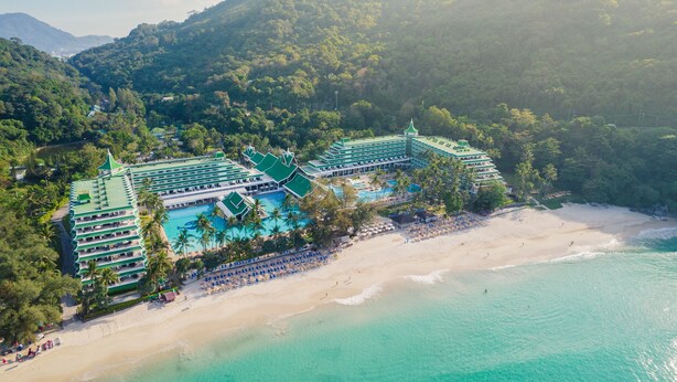 Vista aérea de um resort com vários edifícios com telhados verdes, uma praia e uma piscina.