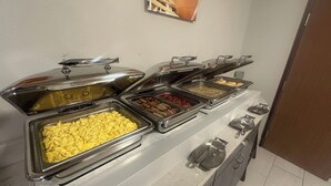 Buffet com diversos alimentos em recipientes de metal, uma máquina de café e uma colher no balcão.